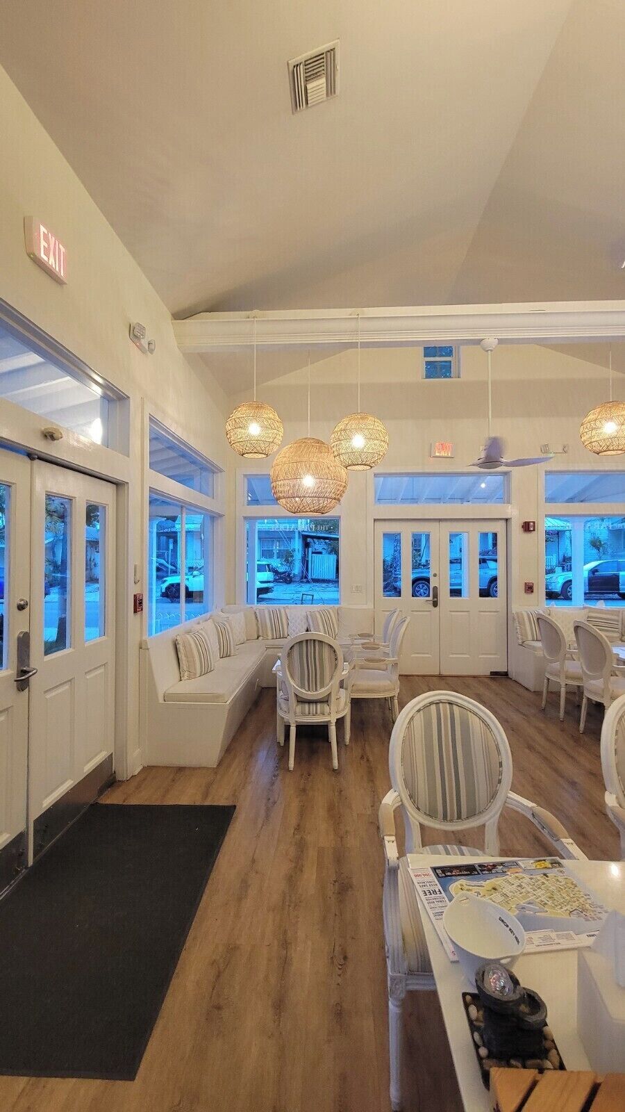 Restaurante com teto alto, luminárias pendentes de vime, mobiliário branco e grandes janelas.