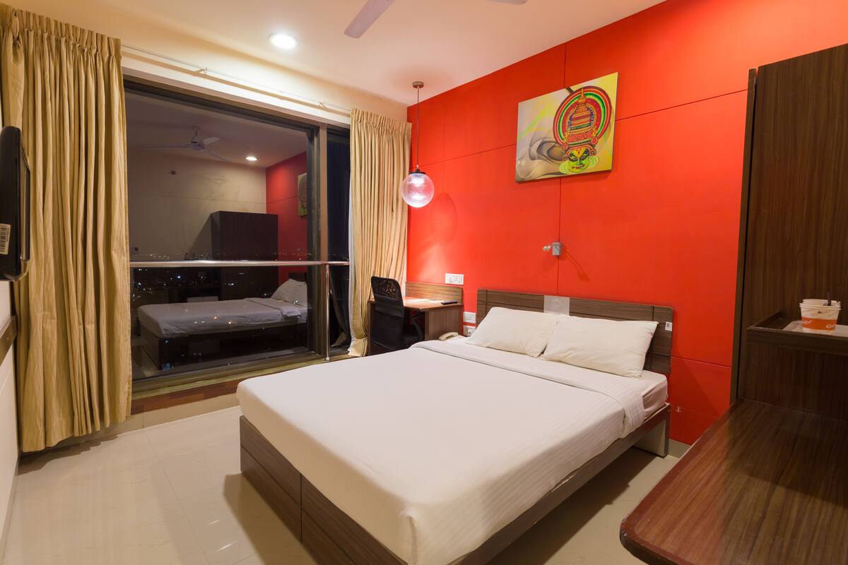 Quarto de hotel com uma cama grande, uma escrivaninha, uma cadeira, uma janela com cortinas e um quadro na parede.