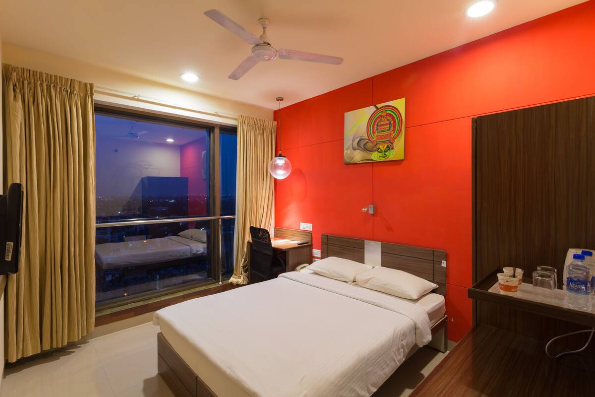 Quarto de hotel com cama, escrivaninha, cadeira, janela com cortinas, ventilador de teto e um quadro na parede.