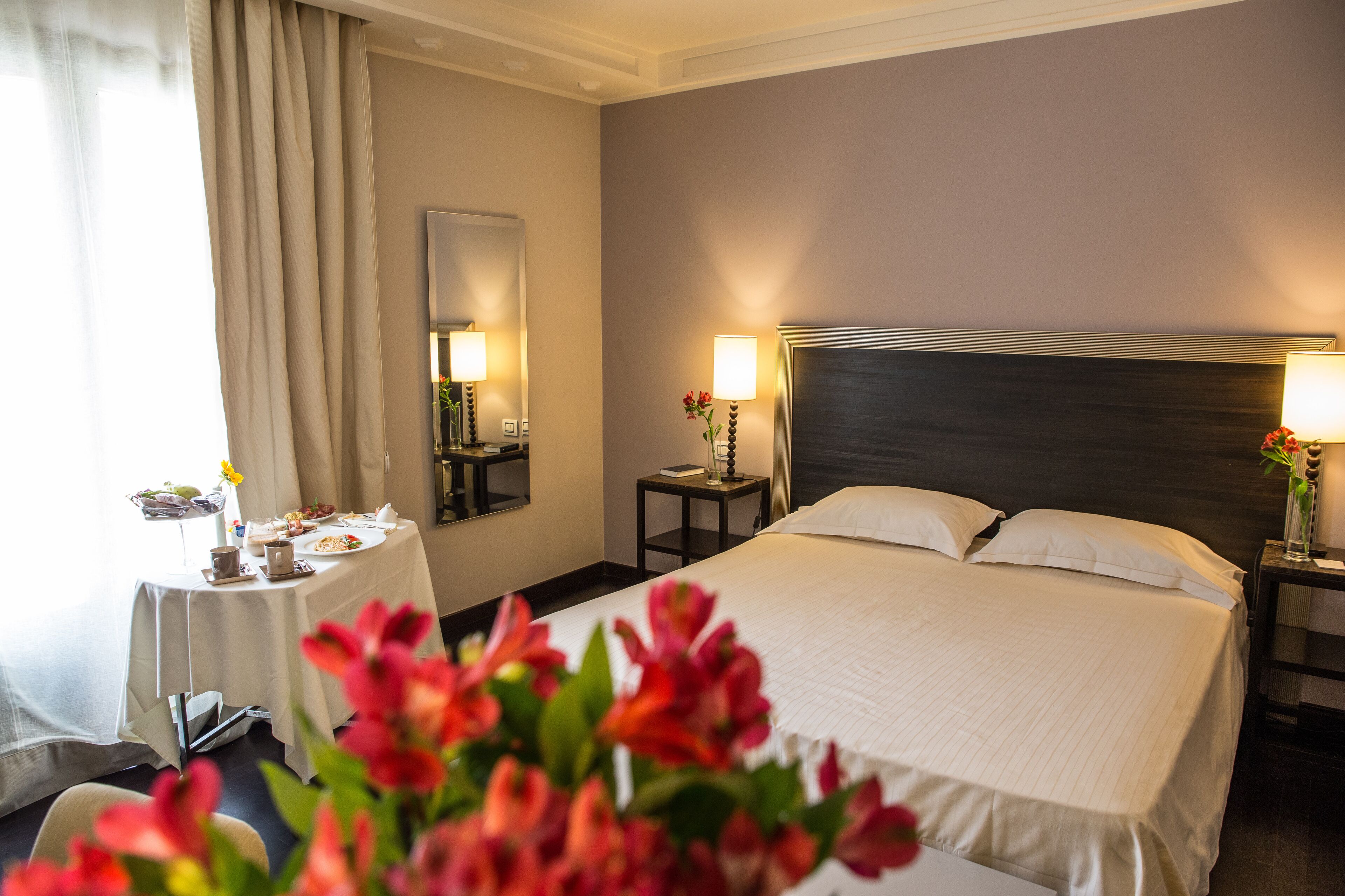 Quarto de hotel com uma cama grande, uma mesa de cabeceira com abajur, uma mesinha com abajur e um espelho na parede.