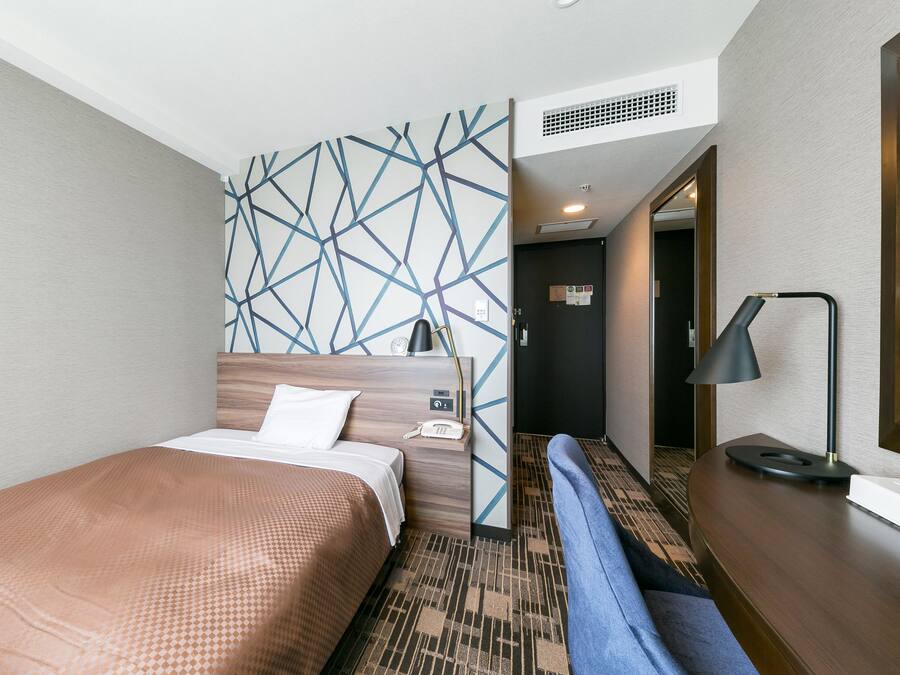 Quarto de hotel com cama, mesa de cabeceira, abajur e parede com padrão.
