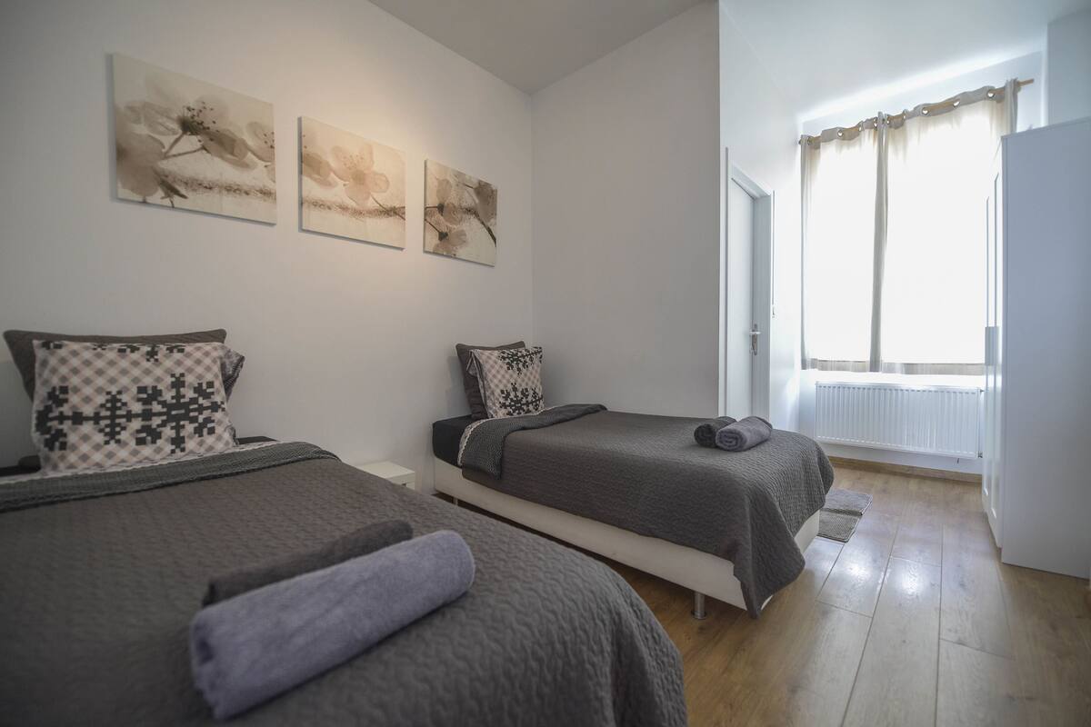 Quarto com duas camas, um radiador e uma janela com cortinas.