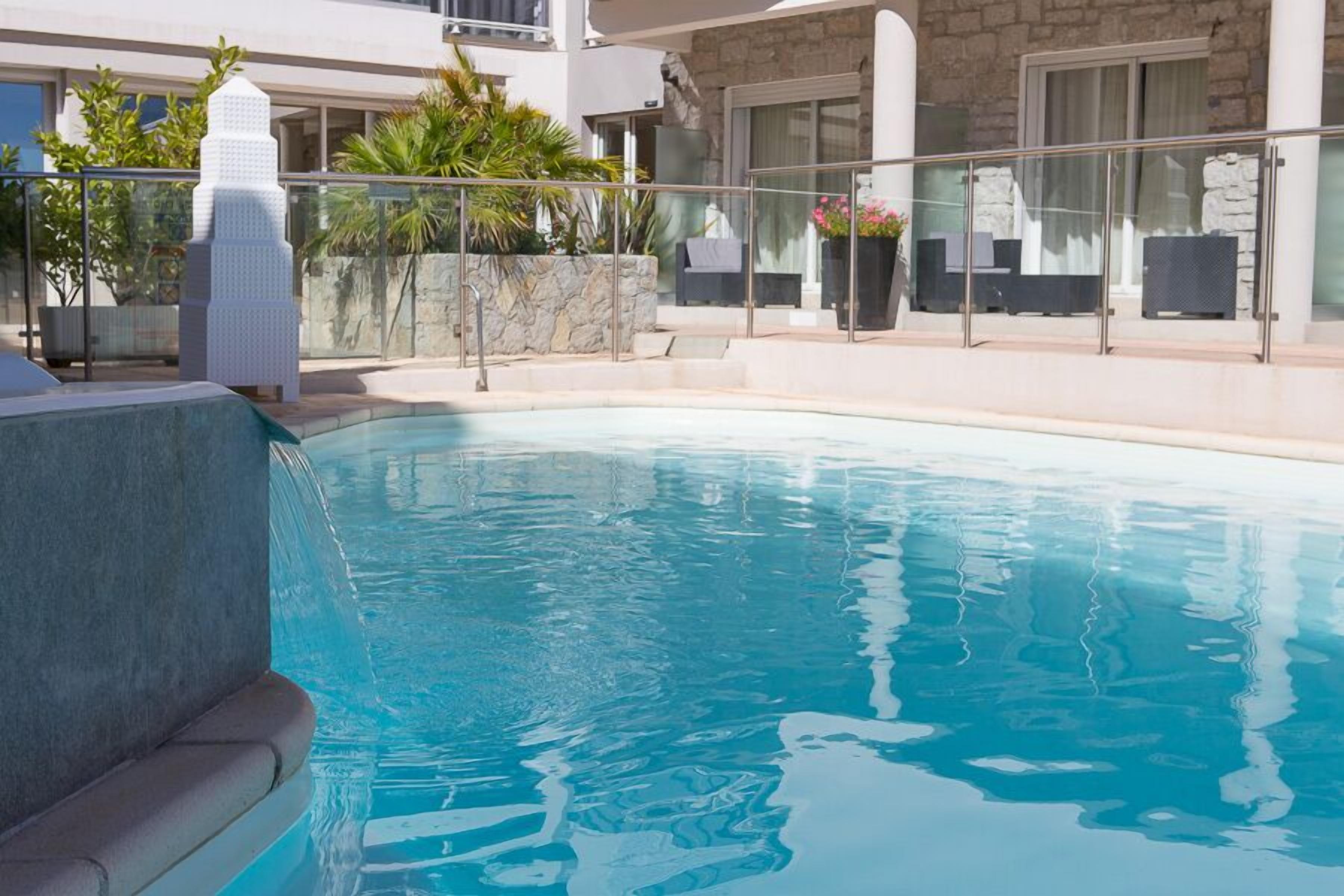 Piscina com guarda-corpo de vidro, parede de pedra e um edifício moderno com varandas.