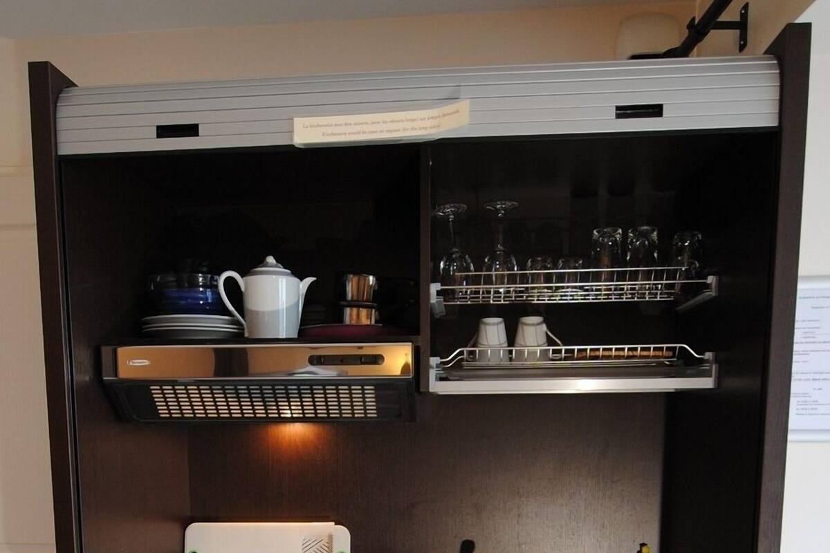 Uma cozinha compacta com forno embutido, pia e bancada com chaleira, xícaras e uma garrafa.