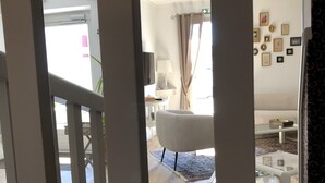 Uma escadaria com carpete estampado que leva a uma sala de estar com uma poltrona branca, uma mesa de centro de vidro e uma parede com quadros.