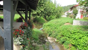 Um pequeno riacho com pedras e vegetação, uma estrutura de madeira com telhado, e um prédio com telhado vermelho ao fundo.