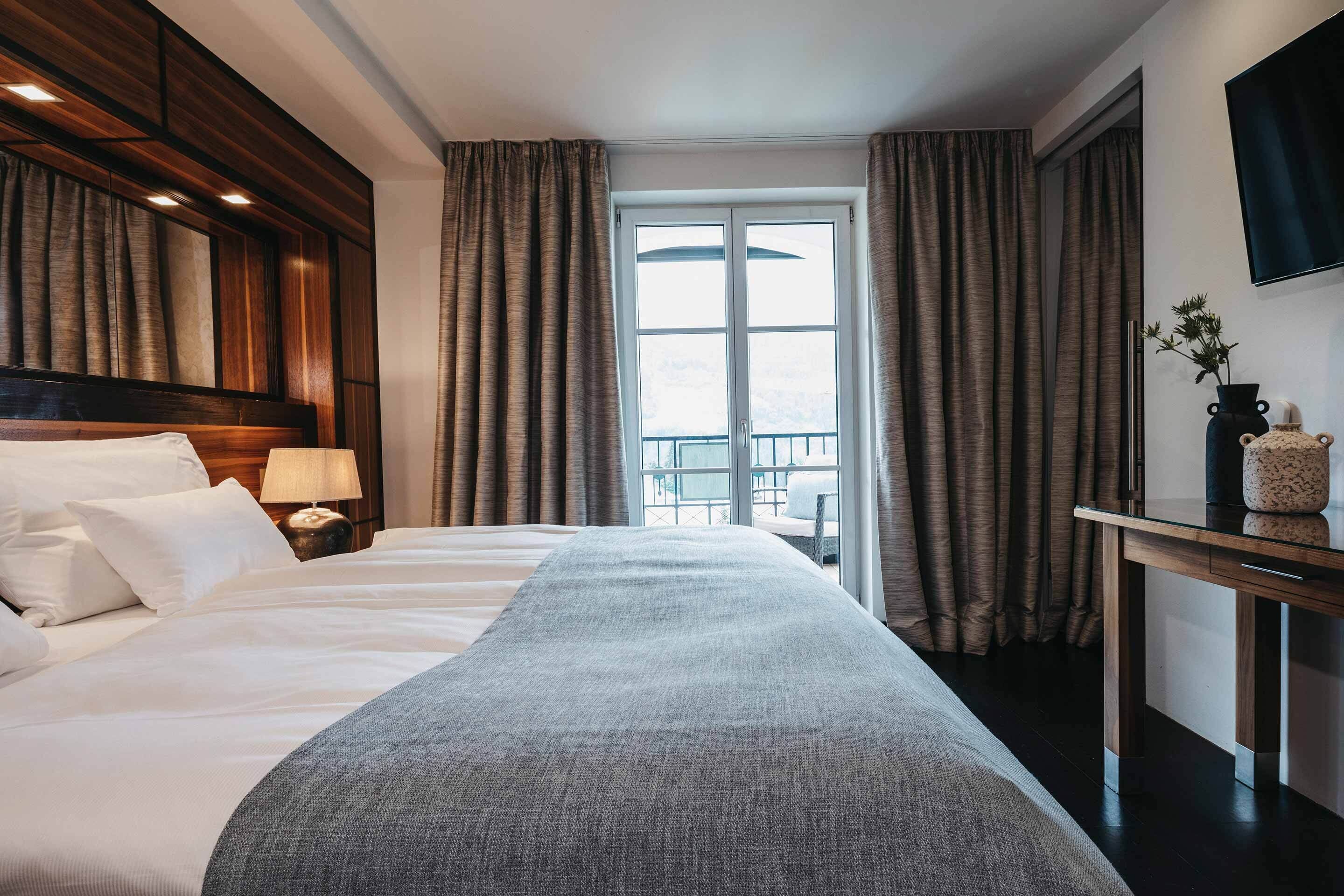 Quarto de hotel com uma cama grande, cabeceira de madeira, mesa de cabeceira com abajur, janela com cortinas e vista para o exterior.