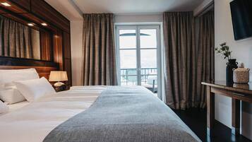 Quarto de hotel com uma cama grande, cabeceira de madeira, mesa de cabeceira com abajur, janela com cortinas e vista para o exterior.