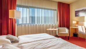 Quarto de hotel com duas camas, uma mesa redonda pequena, uma cadeira, uma escrivaninha e uma janela com cortinas.