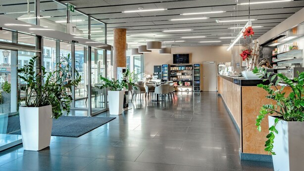 Um hall moderno e bem iluminado, com paredes de vidro, balcão de recepção, área de estar e plantas em vasos.