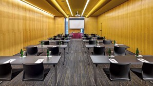 Uma sala de conferência com mesas e cadeiras dispostas em fileiras, uma tela grande na frente e piso coberto por carpete.