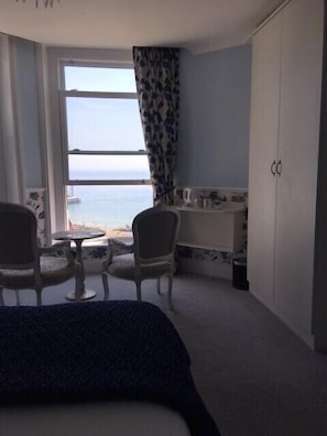 Quarto de hotel com uma grande janela para o mar, uma cama, duas cadeiras, uma mesinha e um guarda-roupa.