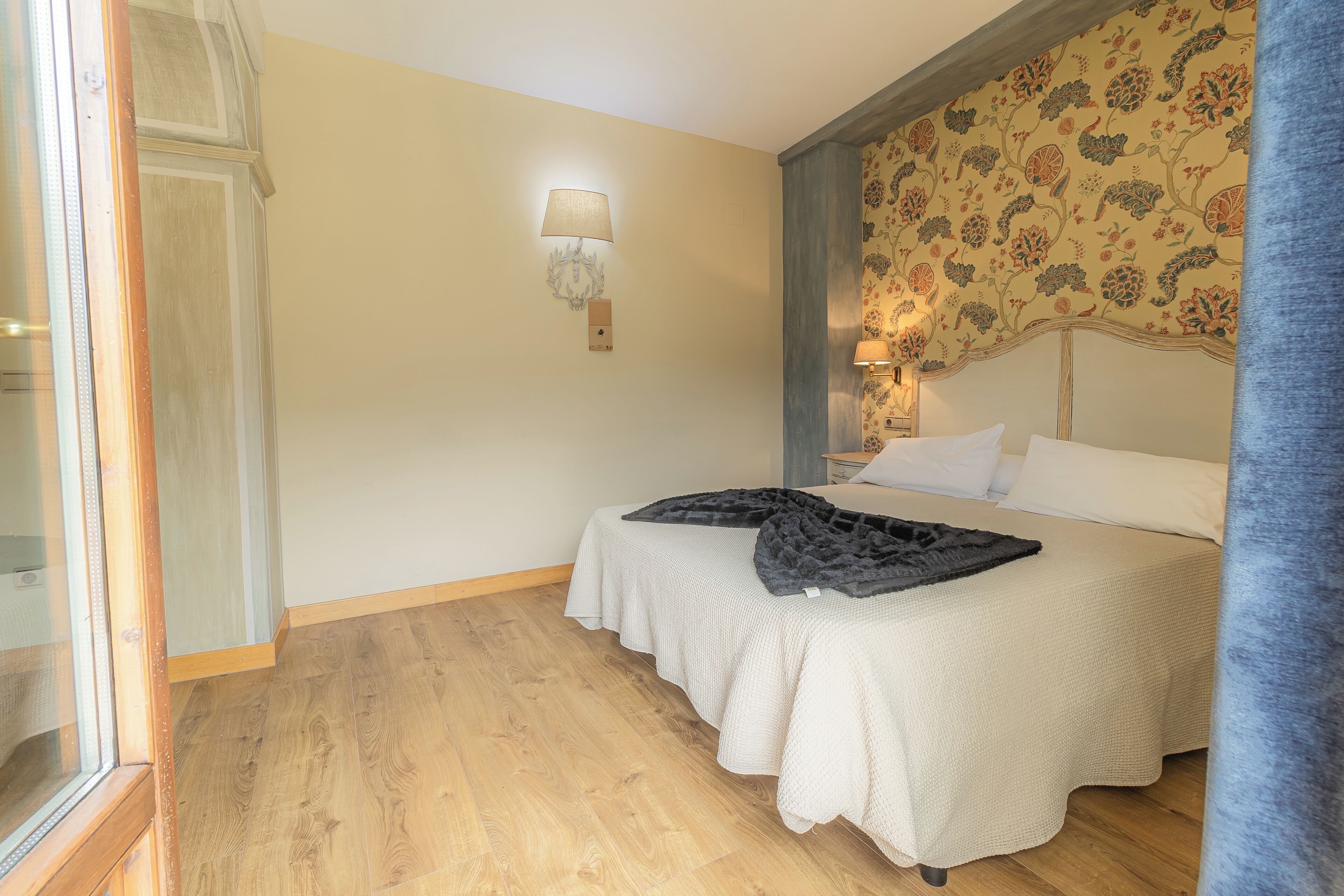 Quarto com cama, mesa de cabeceira, luminária na parede e janela com vista para o exterior.