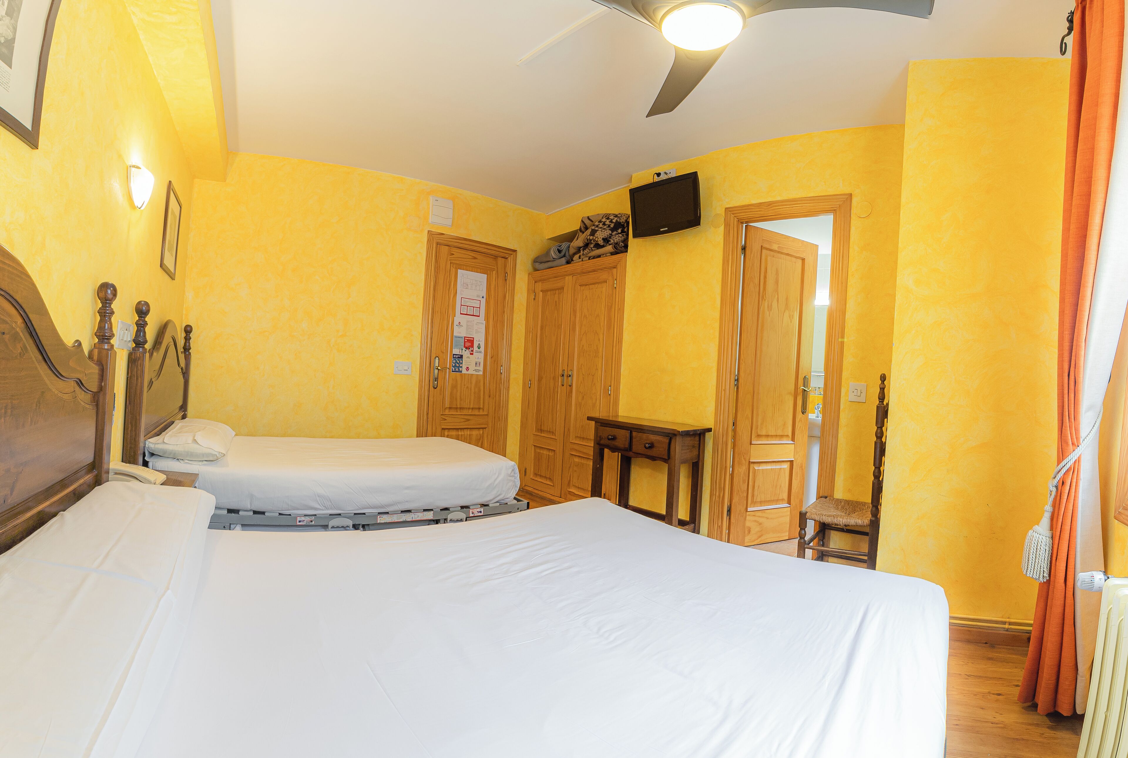 Quarto com duas camas, mobiliário de madeira, ventoinha no teto e televisão.