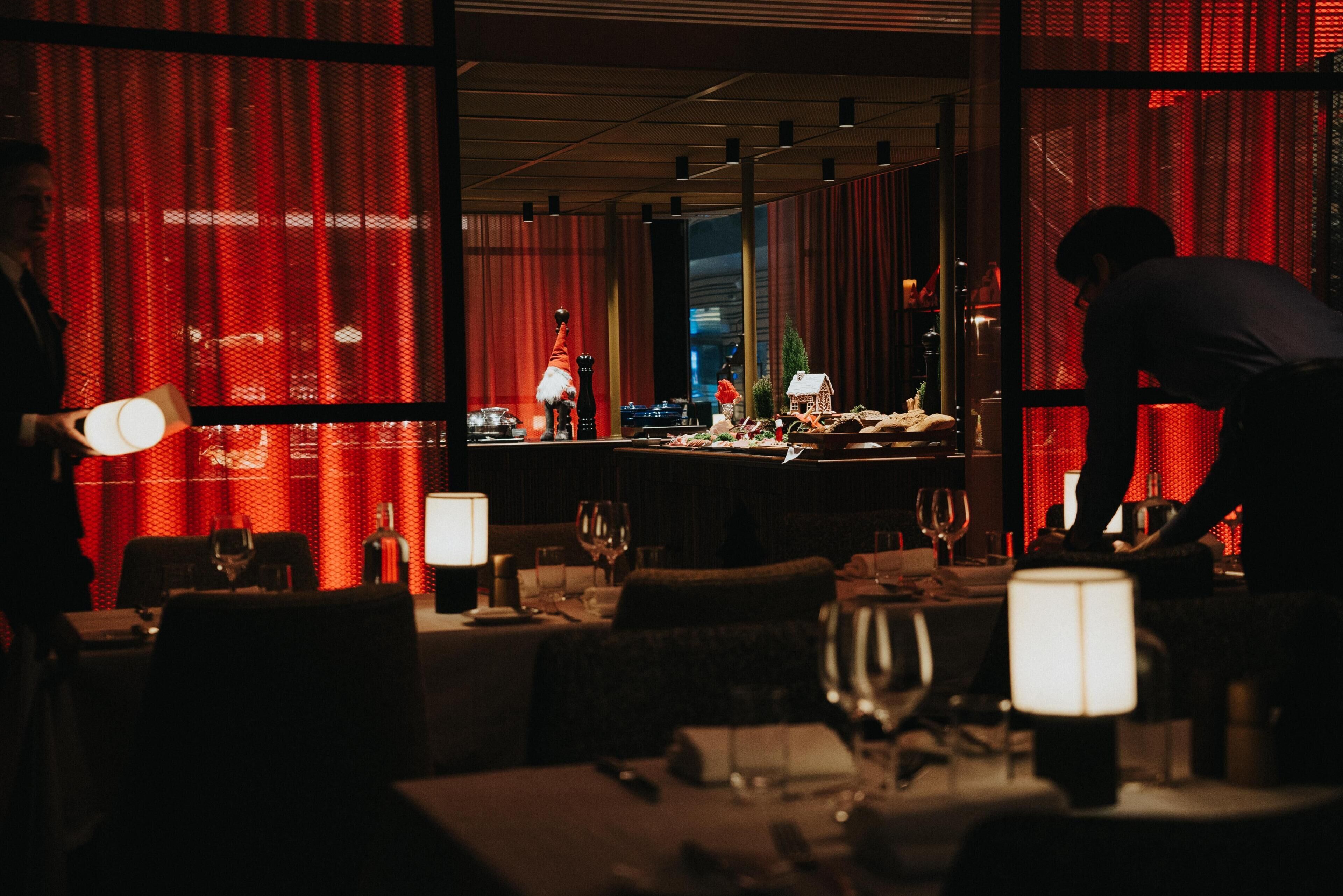 Um restaurante com pouca luz, cortinas vermelhas, mesas postas com copos de vinho e um bartender preparando bebidas.