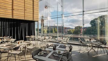 Restaurante moderno com grandes janelas que oferecem vista panorâmica da cidade. Mesas dispostas de forma organizada com louças postas.