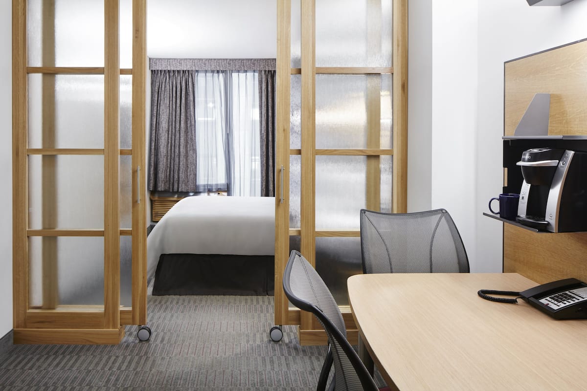 Quarto de hotel com cama, escrivaninha, cadeira, telefone, máquina de café e janela com cortinas.