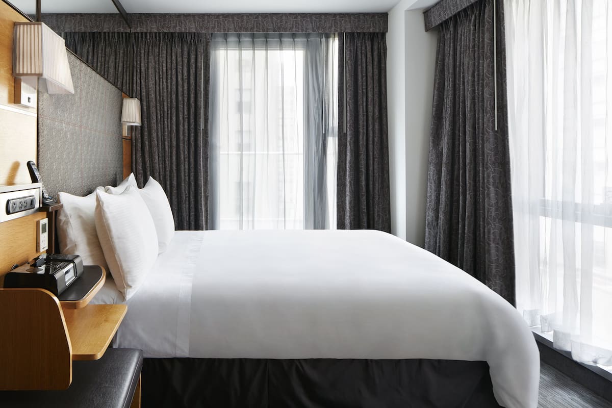 Quarto de hotel com uma cama grande, uma mesa de cabeceira com luminária, uma escrivaninha com telefone e uma janela com cortinas.