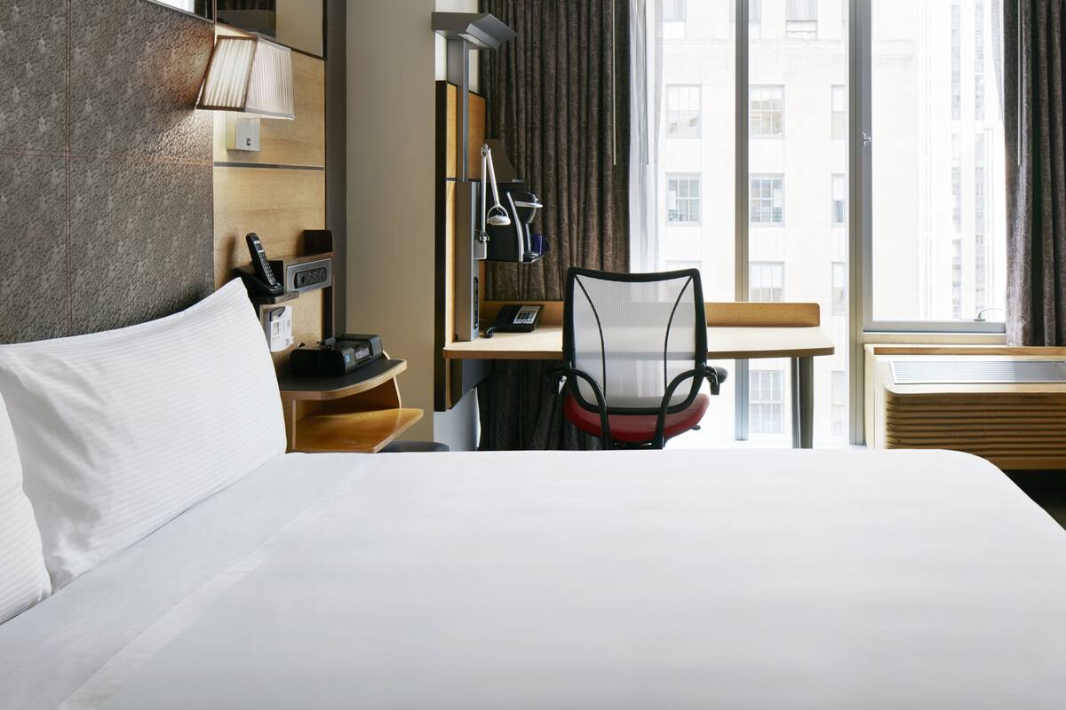 Quarto de hotel com uma cama grande, uma mesa com cadeira, uma luminária, um telefone e uma janela com cortinas.