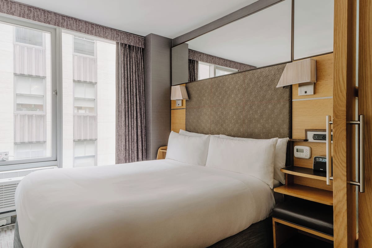 Quarto de hotel com uma cama grande, mesa de cabeceira com abajur, espelho e relógio digital.
