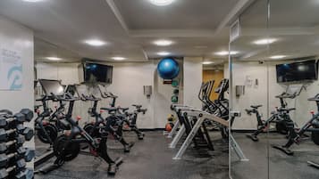 Uma academia com várias bicicletas de exercício, um espelho grande na parede e uma TV fixada na parede.