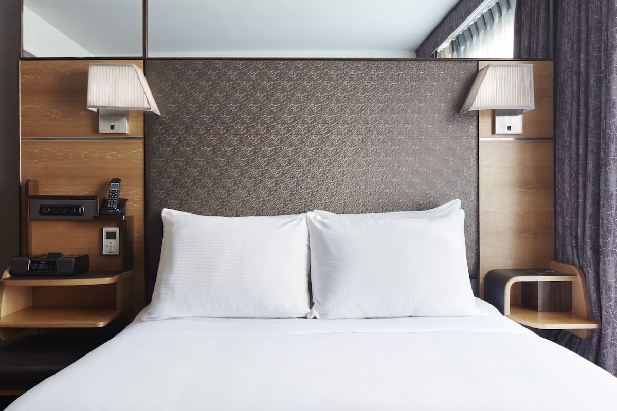 Quarto de hotel moderno com uma cama grande, uma mesa de cabeceira com telefone e relógio, e cabeceira com padrão texturizado.