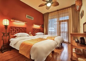 Quarto de hotel com duas camas, cabeceiras de madeira, um ventilador de teto, mesa de cabeceira, abajur e vista do exterior por grandes janelas.