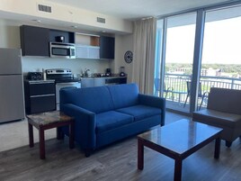 Uma sala de estar moderna com um sofá azul, uma mesa de centro de madeira e uma cozinha com micro-ondas, forno e máquina de café.