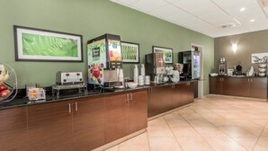 Uma área de recepção de hotel com balcão, máquinas de café, dispensador de suco de fruta e geladeira.