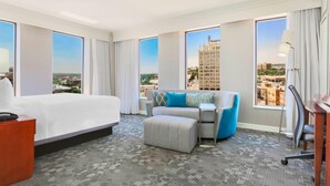 Quarto de hotel com uma cama grande, sofá, banquinho, escrivaninha e uma cadeira. Janelas amplas com vista para a cidade.