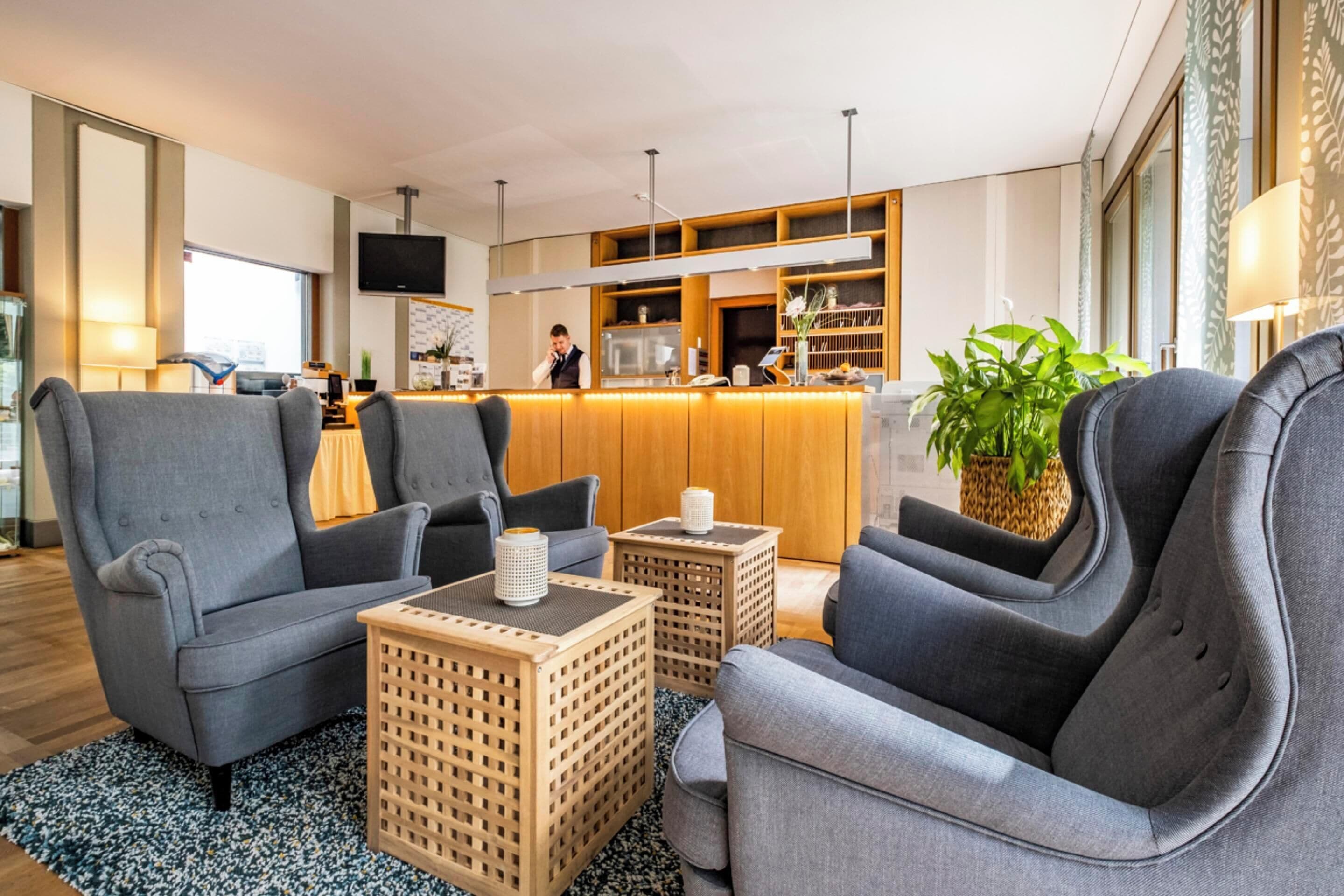 Um saguão de hotel moderno com sofás cinzas, um balcão de recepção de madeira e uma grande planta em vaso.