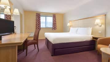 Quarto de hotel com cama, escrivaninha, cadeira, televisão e janela com cortinas.