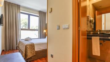 Quarto de hotel com cama, um pequeno sofá, janela com cortinas, banheiro com espelho, toalha pendurada no porta-toalhas e telefone fixado na parede.