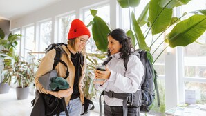 Duas pessoas com mochilas em um quarto com janelas amplas e plantas em vasos.