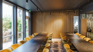 Sala de conferências com mesas compridas e cadeiras amarelas, janelas amplas e painel de ripas de madeira.
