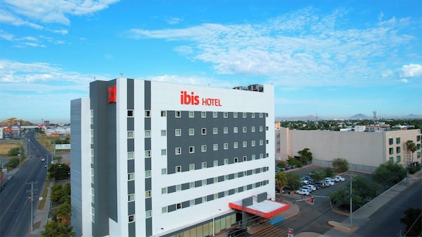 Um edifício de hotel moderno com um sinal de destaque "ibis HOTEL", vários andares com padrões de janelas uniformes e uma área de estacionamento com diversos carros.