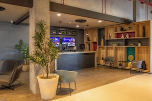 Uma área de recepção moderna com balcão de madeira, duas cadeiras, uma grande planta em vaso e uma TV fixada na parede.