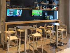 Uma área de bar com diversas opções de assento, incluindo mesas e cadeiras, e uma tela de televisão exibindo conteúdo esportivo.