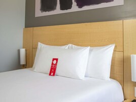 Cama bem arrumada com roupa de cama e almofadas brancas, cabeceira de madeira e luminária na parede.