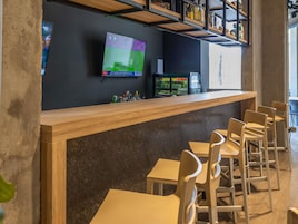 Uma área de bar com balcão, cadeiras, tela de televisão e prateleiras com garrafas.
