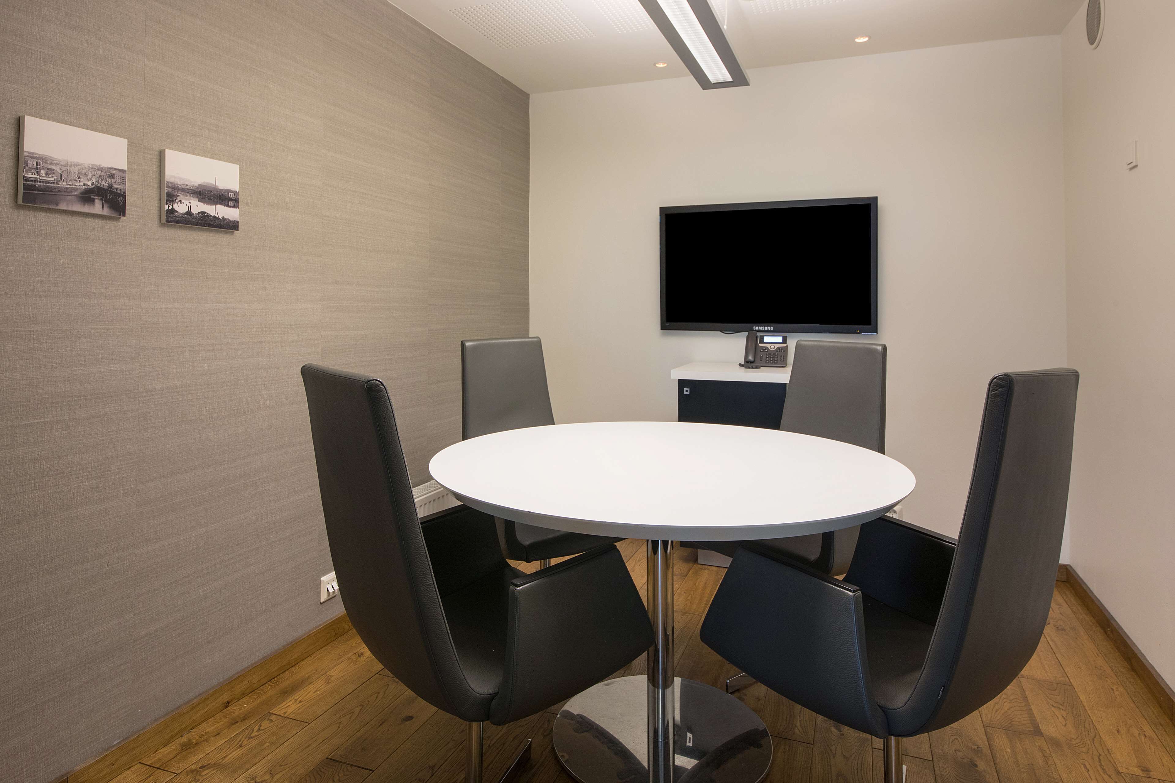 Uma sala de conferências com uma mesa redonda, quatro cadeiras pretas, uma televisão de tela plana e duas imagens emolduradas na parede.