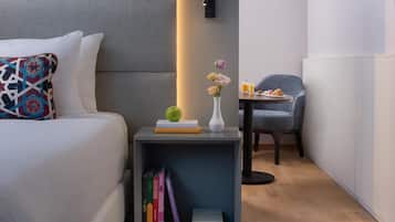 Quarto de hotel moderno com cama bem arrumada, mesa de cabeceira com livros e um vaso de flores, pequena mesa com itens de café da manhã e uma cadeira.