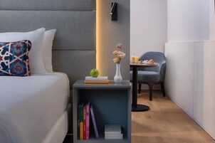 Quarto de hotel moderno com cama bem arrumada, mesa de cabeceira com livros e um vaso de flores, pequena mesa com itens de café da manhã e uma cadeira.
