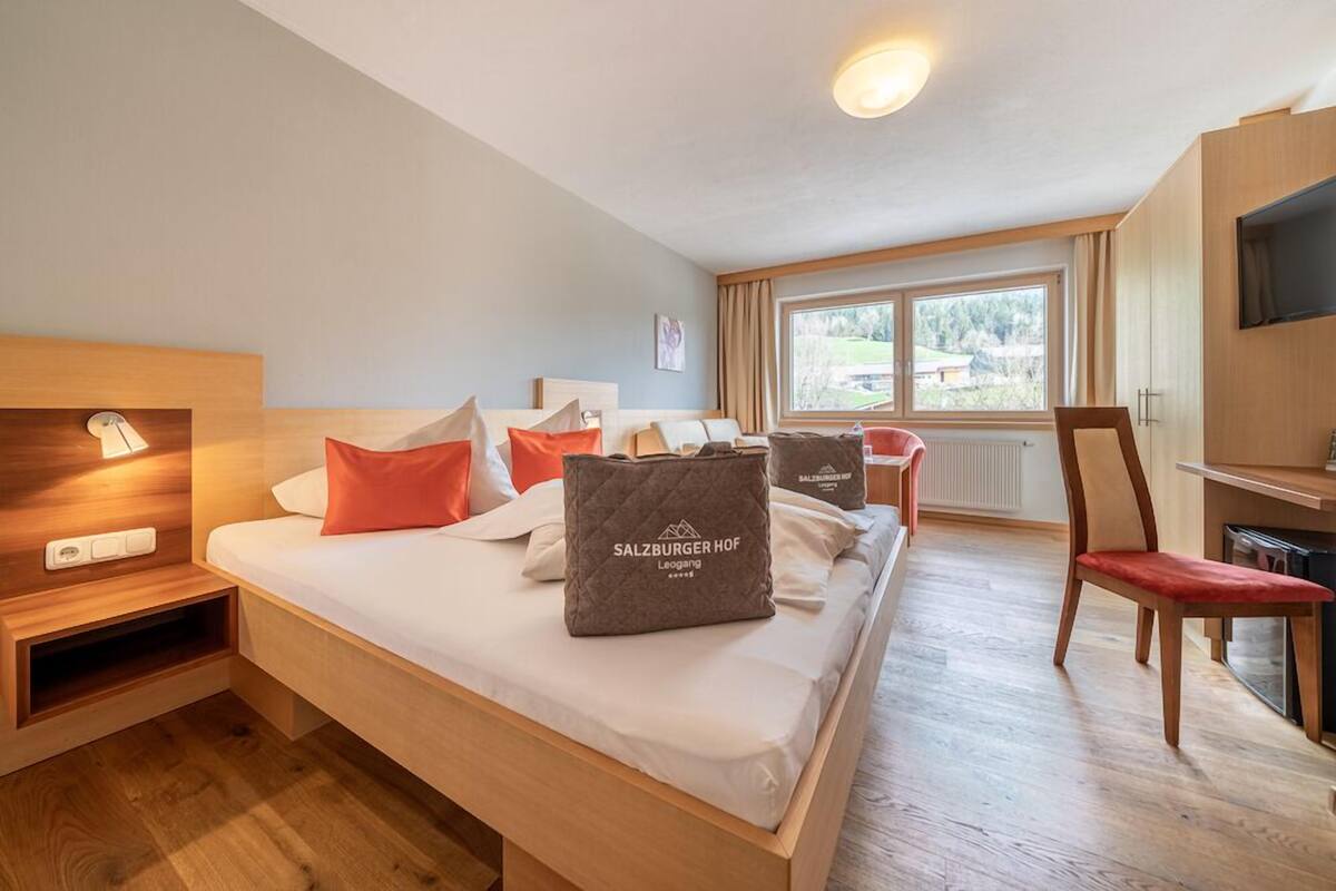 Quarto de hotel com cama, escrivaninha, cadeira e televisão.
