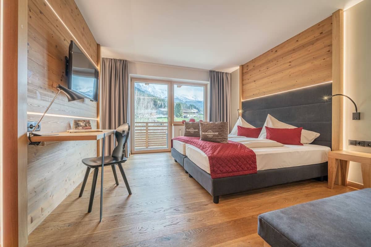 Quarto de hotel moderno com decoração em madeira, cama com roupa de cama vermelha e branca, escrivaninha com cadeira e uma grande janela com vista para as montanhas.