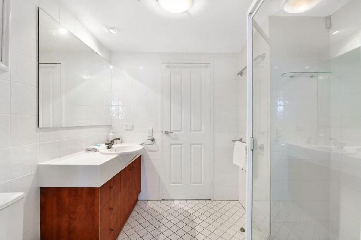 Um banheiro moderno com piso de azulejos brancos, toucador de madeira, espelho grande, box de vidro e porta branca.