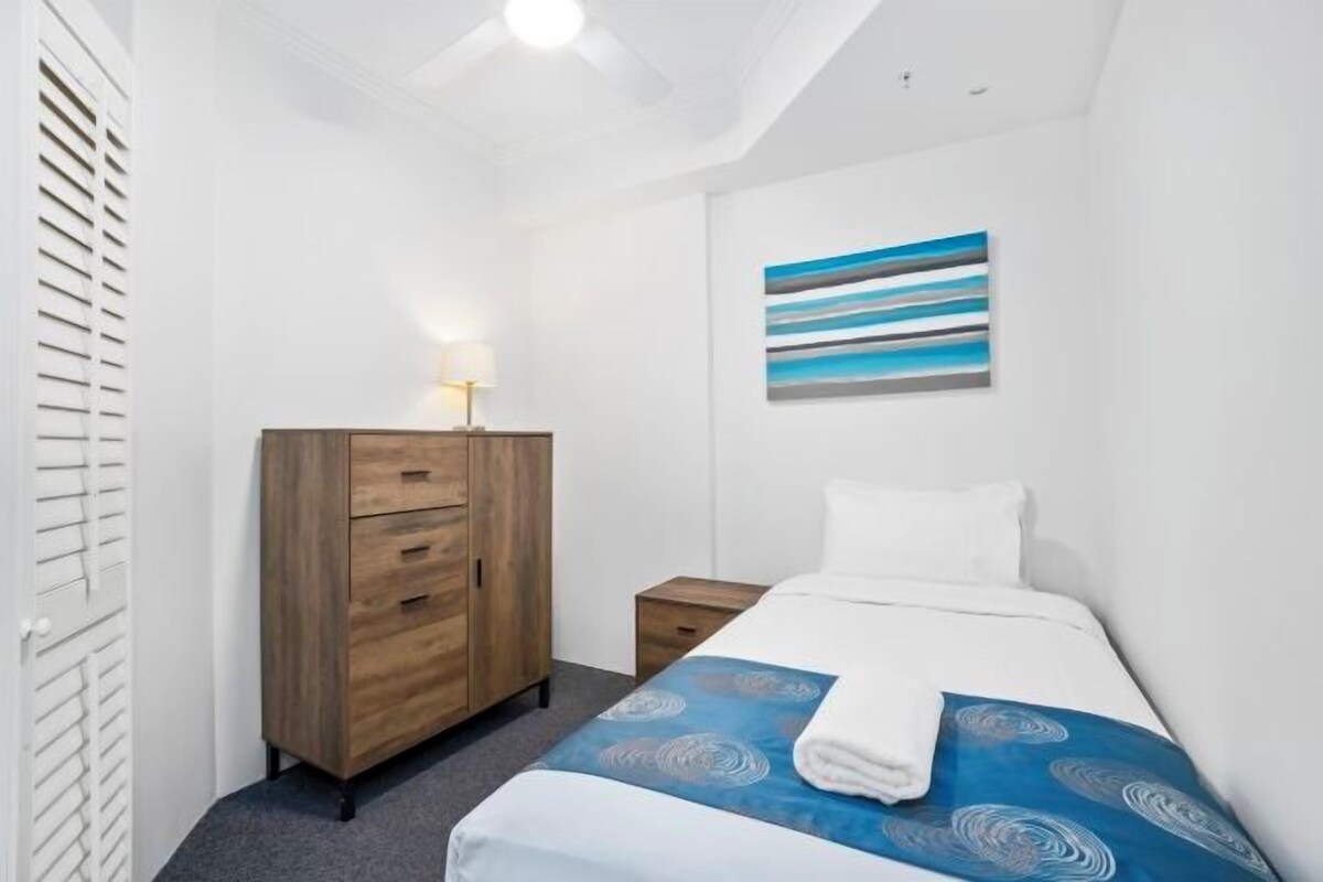 Quarto de hotel com cama, guarda-roupa de madeira, mesa de cabeceira, luminária e um quadro na parede.