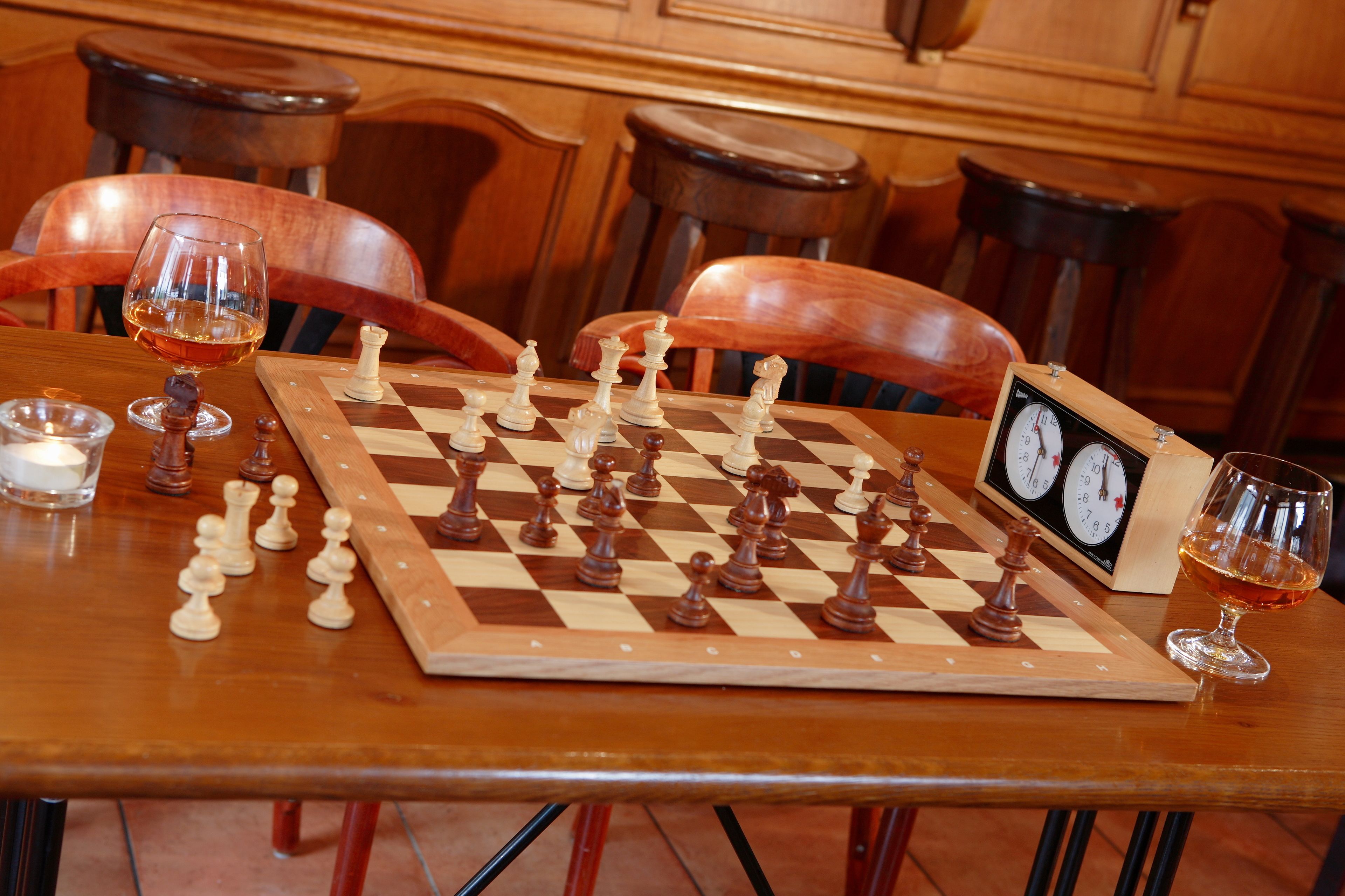 Uma mesa de madeira com um tabuleiro de xadrez, dois copos com líquido âmbar e um relógio.