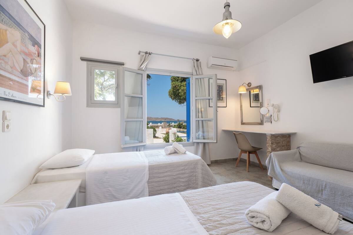 Quarto de hotel com duas camas, uma escrivaninha, uma cadeira, televisão e vista para o mar.