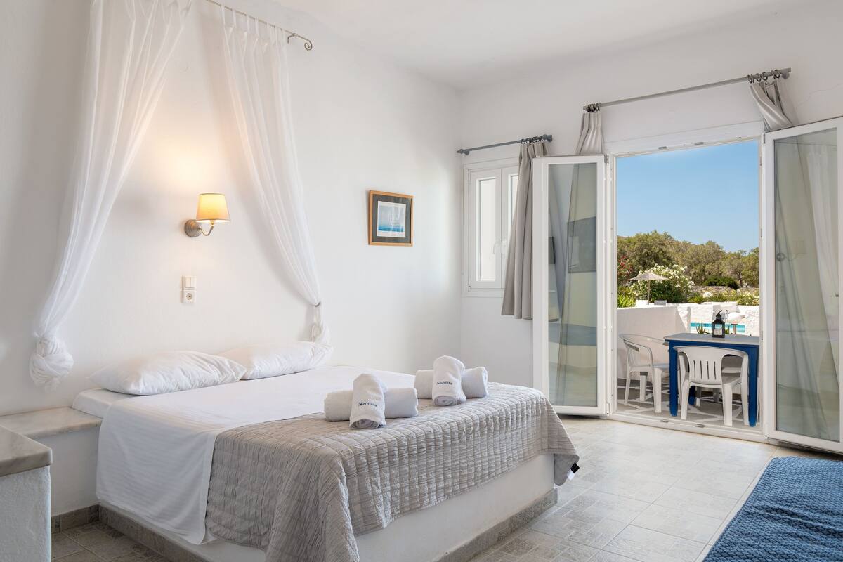 Quarto com cama, mesa de cabeceira, luminária e vista para a varanda.